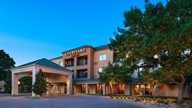 Um hotel Courtyard Marriott com entrada coberta, vários andares e área ajardinada com árvores e arbustos.