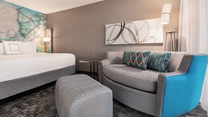 Quarto de hotel com cama, sofá cinza com detalhe azul, mesinha e parede com arte abstrata.
