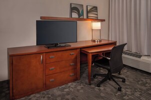 Quarto de hotel com secretária de madeira, televisão de tela plana, luminária, cadeira e uma grande janela com cortinas.