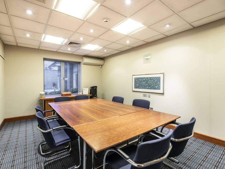 Uma sala de conferências com uma grande mesa de madeira, cadeiras pretas, uma janela com persianas, um quadro na parede e um aparelho de ar condicionado.