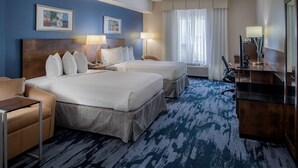 Quarto de hotel com uma cama grande, uma cama menor, uma escrivaninha, uma cadeira e uma luminária de mesa.