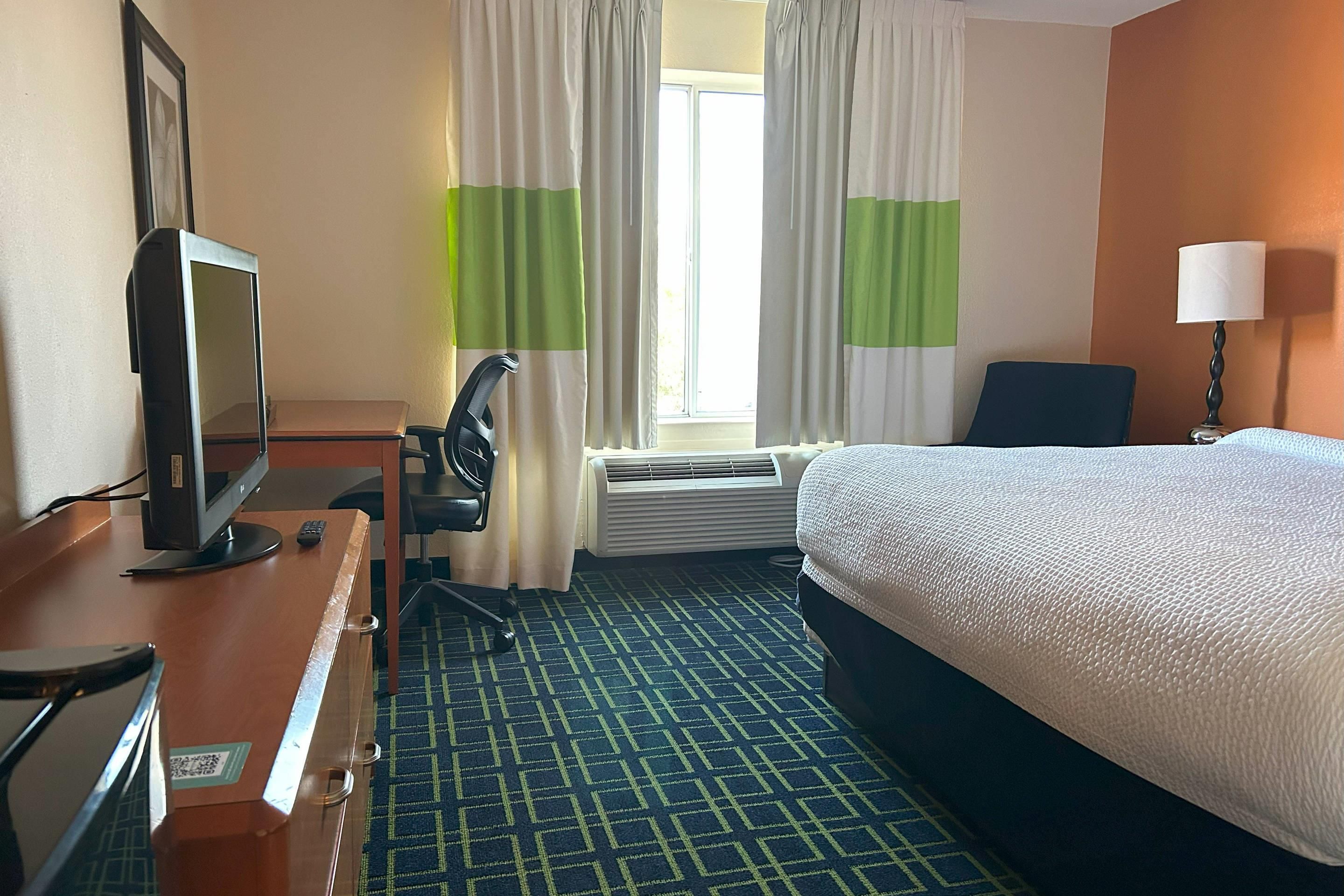 Quarto de hotel com cama, escrivaninha, cadeira, televisão, aparelho de ar condicionado e cortinas.