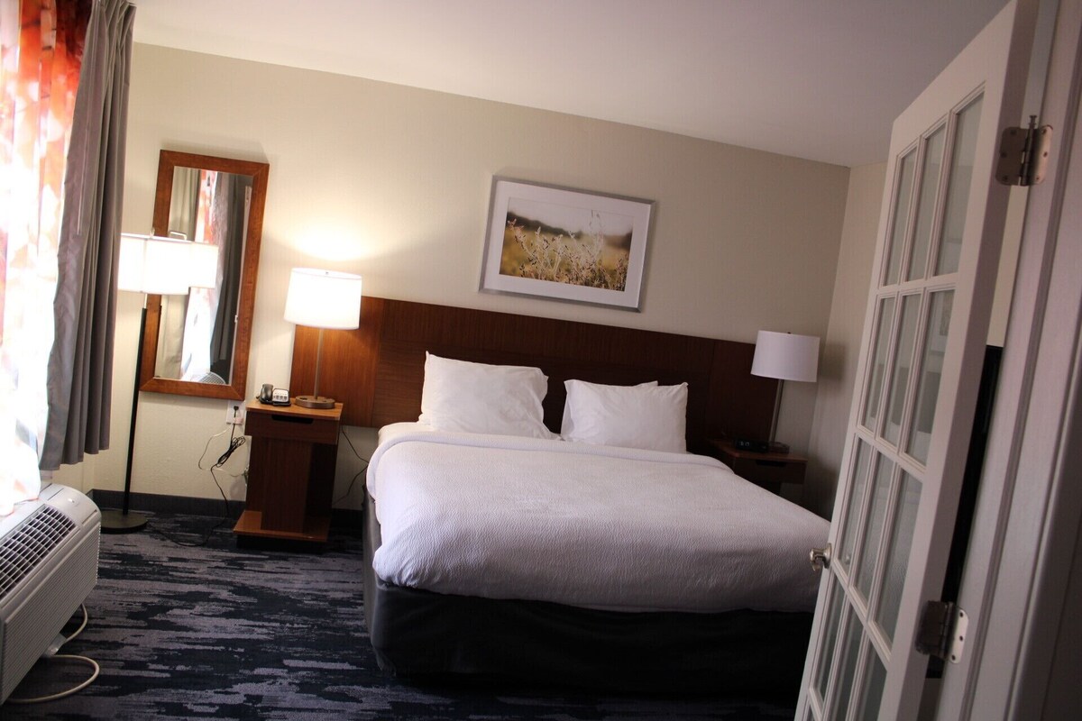 Quarto de hotel com uma cama grande, uma mesa de cabeceira com luminária, um quadro na parede e uma janela com cortinas.