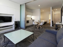 Quarto de hotel moderno com televisão de tela plana, uma kitchenette compacta, área de jantar com mesa e cadeiras de vidro e um sofá.