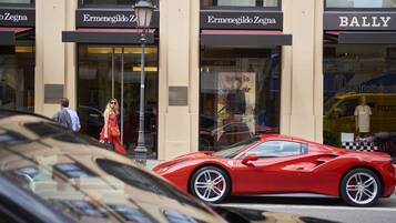 Um carro esportivo vermelho estacionado em frente a um prédio com lojas, incluindo Ermenegildo Zegna e BALLY.