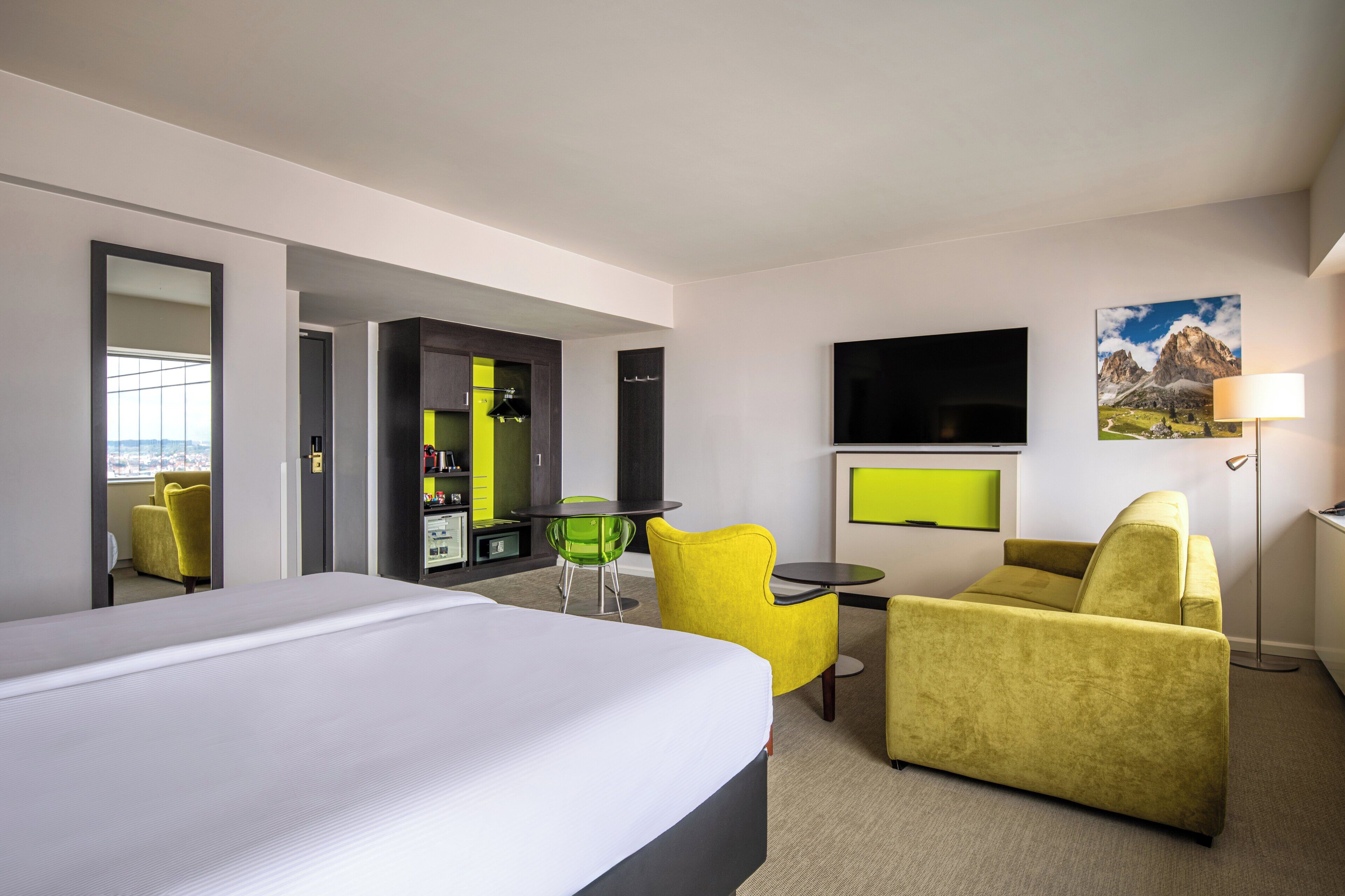 Quarto de hotel com uma cama grande, uma escrivaninha com cadeira, um sofá, uma TV e um abajur.