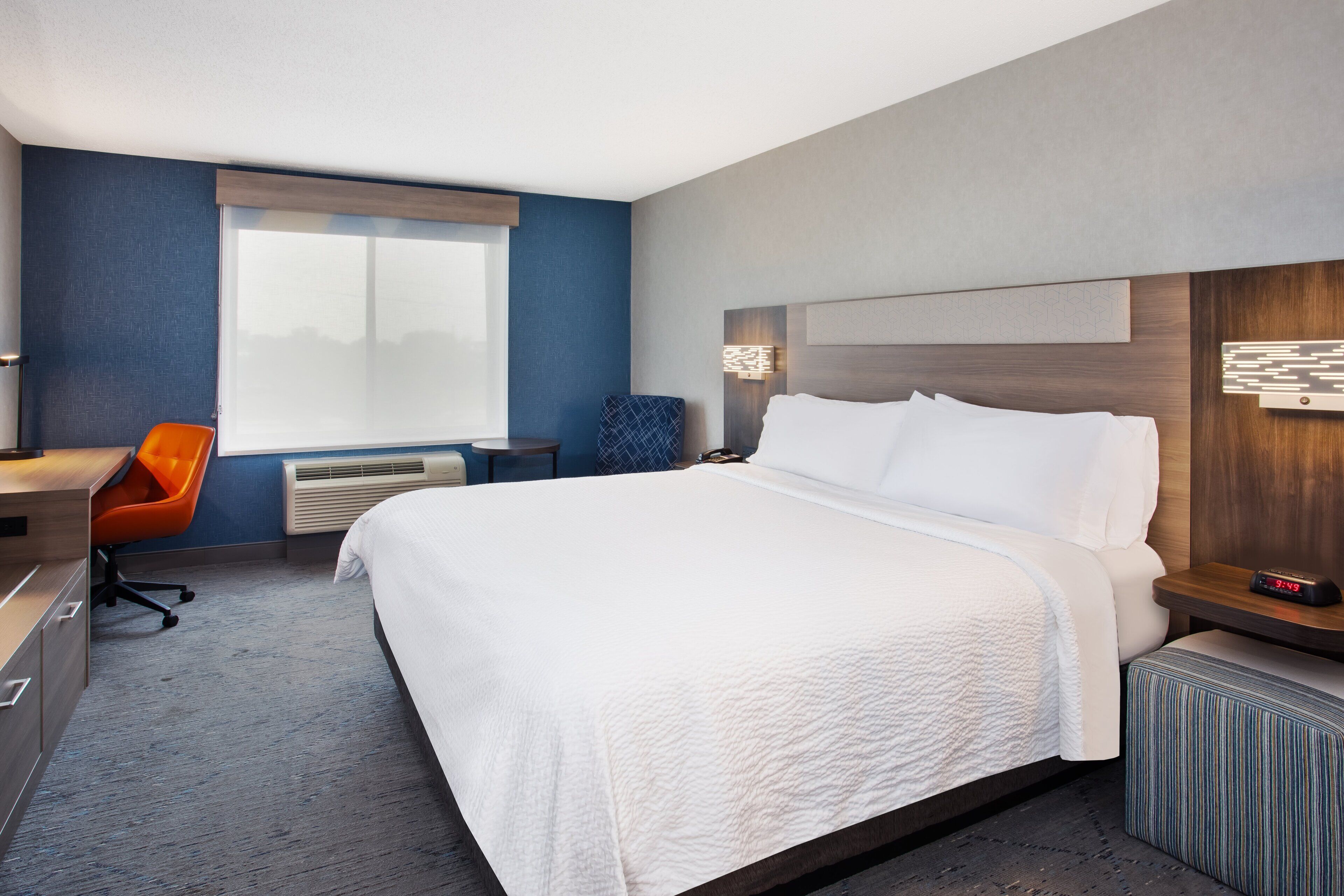 Quarto de hotel moderno com uma cama grande, uma escrivaninha, uma cadeira laranja e uma janela com persianas.