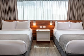 Quarto de hotel com duas camas, cabeceira de madeira, abajures e uma janela com cortinas.