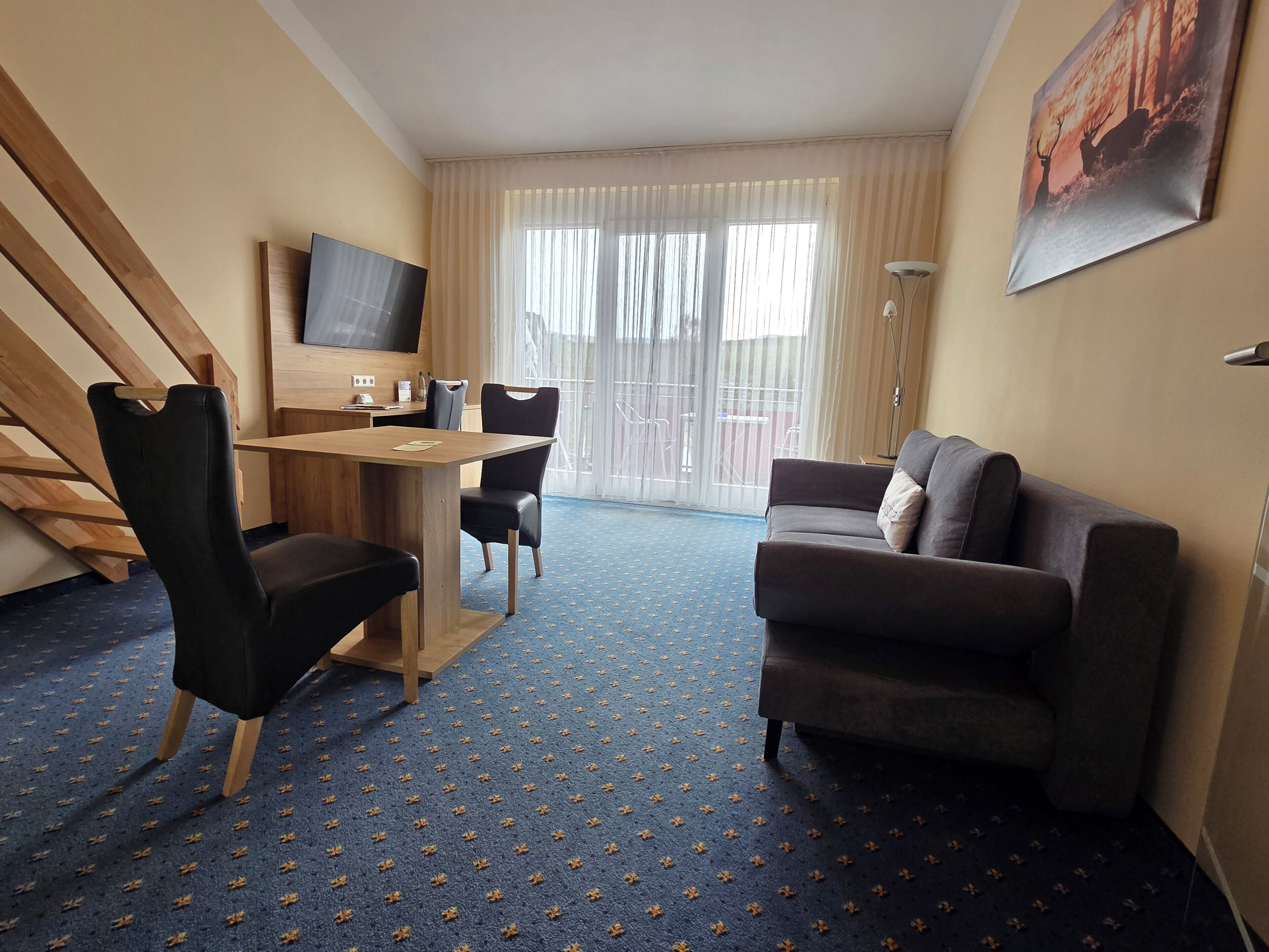 Quarto de hotel com sofá, escrivaninha com cadeira, televisão e janela grande com cortinas.