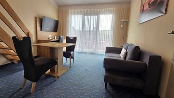 Quarto de hotel com sofá, escrivaninha com cadeira, televisão e janela grande com cortinas.