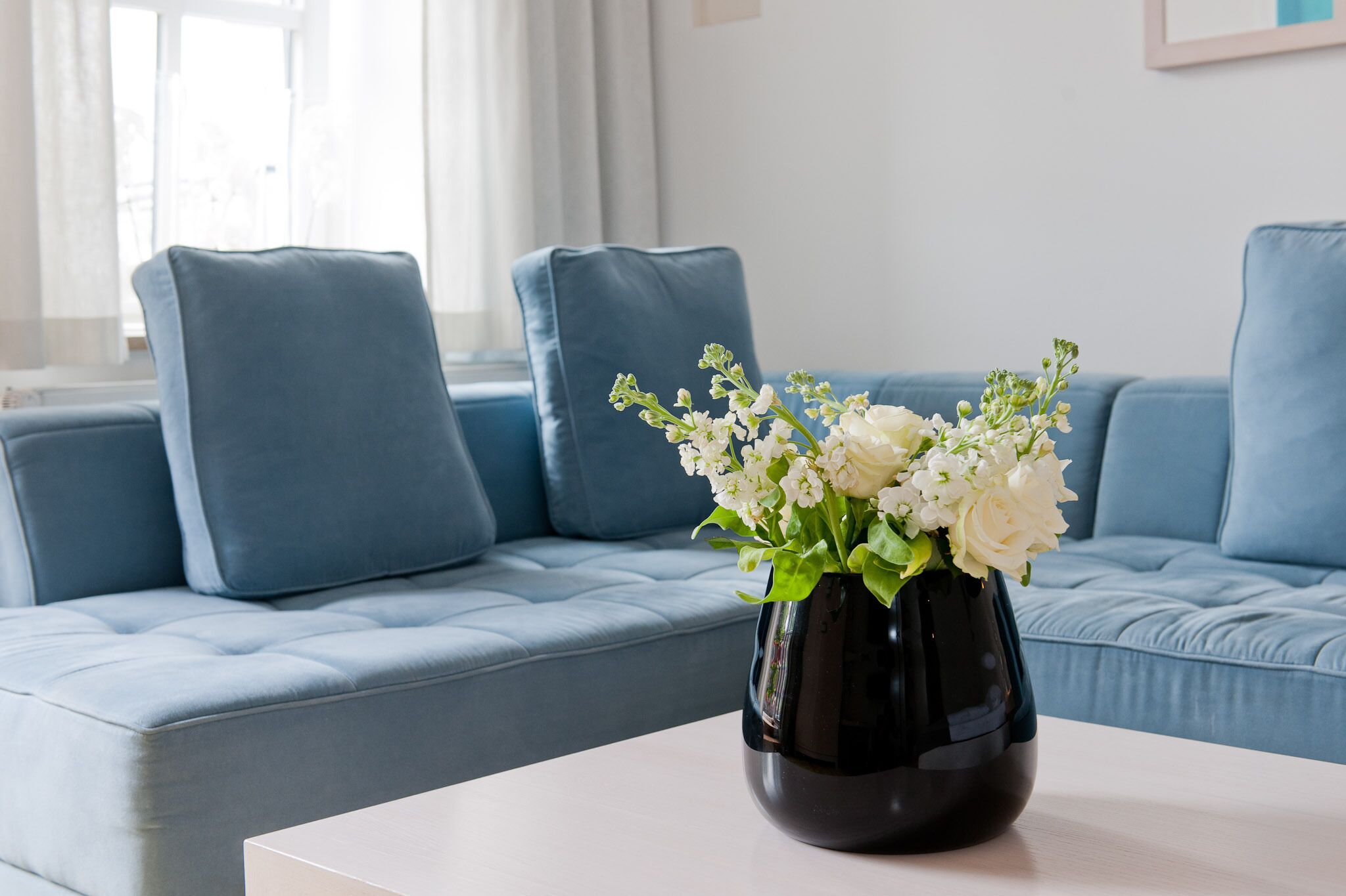 Uma sala de estar moderna com um sofá azul claro e um vaso preto com flores brancas sobre uma mesa branca.