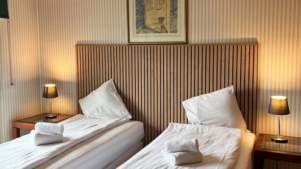 Quarto de hotel com duas camas de solteiro, cada uma com almofada e toalha enrolada, cabeceira de madeira, quadro na parede e duas luminárias de cabeceira.