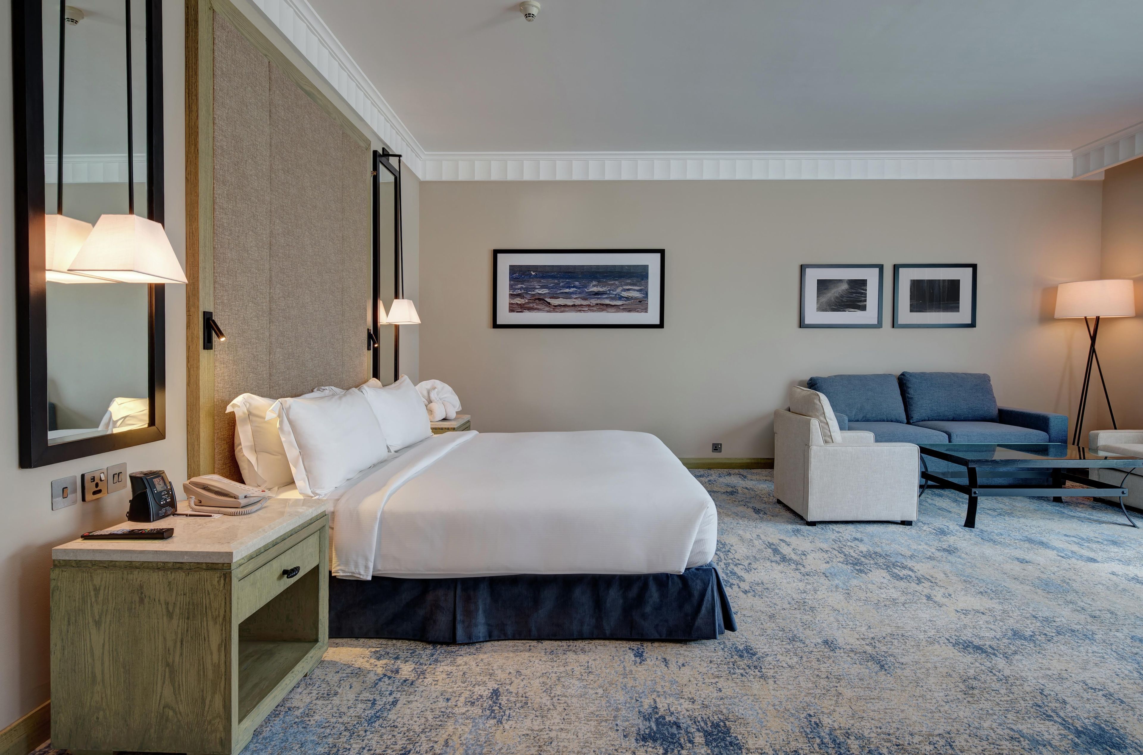 Quarto de hotel com cama, abajures, escrivaninha com telefone, cadeira e duas fotos emolduradas na parede.