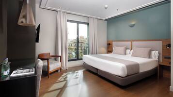 Quarto de hotel moderno com uma cama grande, uma escrivaninha com computador, uma cadeira e vista para a cidade através de uma janela ampla.
