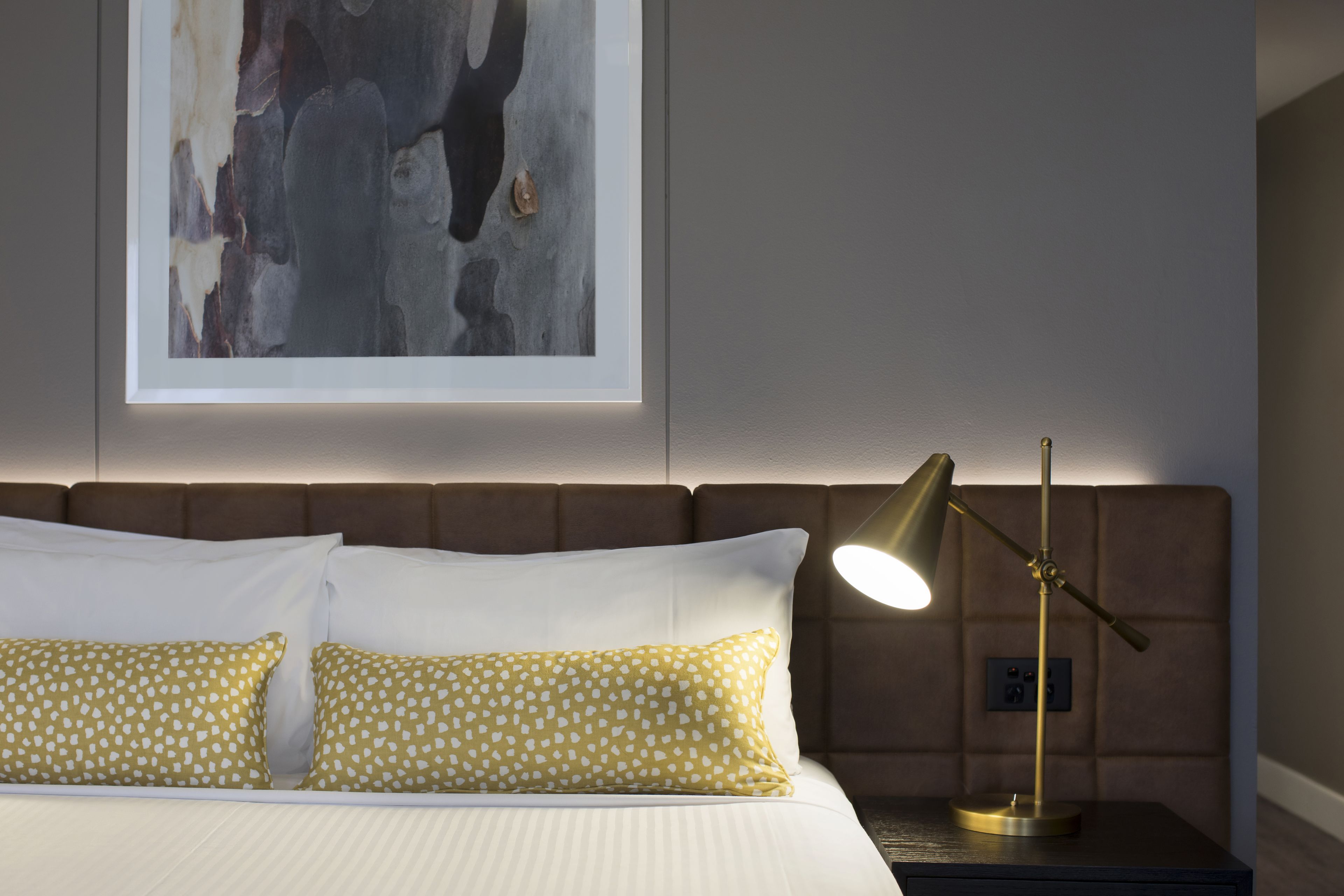 Cama bem arrumada com almofadas brancas e amarelas, abajur e um quadro abstrato na parede.
