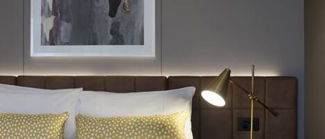 Cama bem arrumada com almofadas brancas e amarelas, abajur e um quadro abstrato na parede.