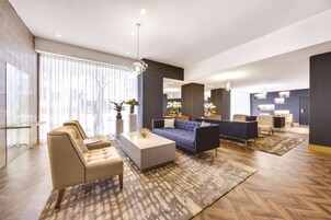 Uma sala de estar moderna com um sofá, poltronas e uma mesa de centro. Há grandes janelas com cortinas leves, um lustre e um espelho na parede.