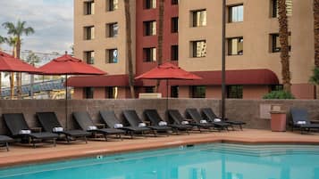 Uma área à beira da piscina com espreguiçadeiras e guarda-sóis, uma piscina límpida e um edifício com várias janelas e varandas.