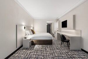 Quarto de hotel com cama, mesa de cabeceira, escrivaninha, cadeira e televisão na parede.