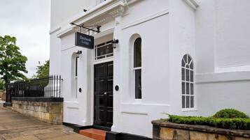 Um edifício branco com porta e janelas pretas, uma placa com a inscrição 'Elmbank Lodge' e um muro de pedra com um pequeno jardim.
