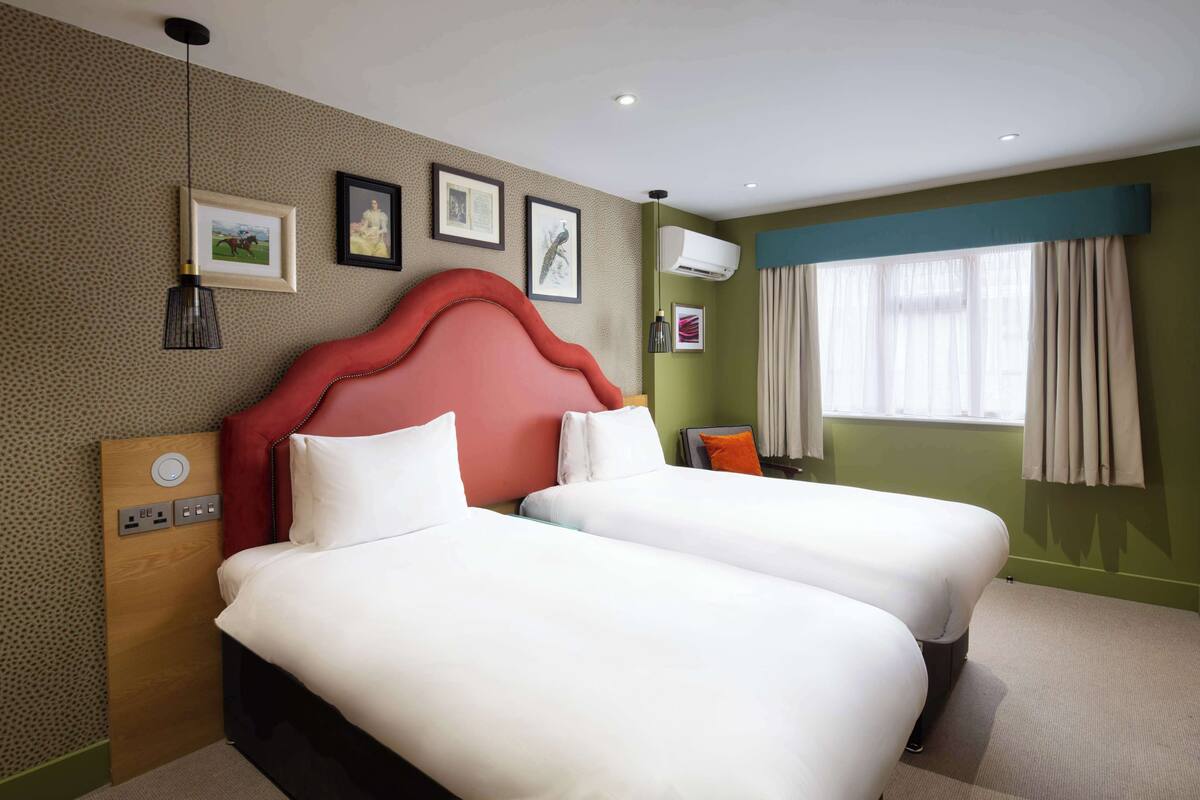 Quarto de hotel com duas camas, cabeceira vermelha, janela com cortinas e ar condicionado fixado na parede.