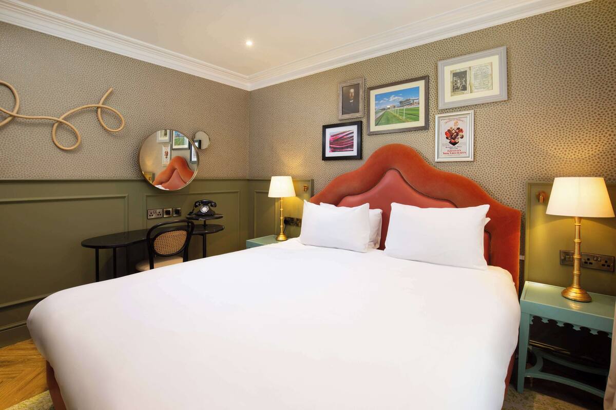 Quarto de hotel com uma cama grande, duas mesas de cabeceira com abajures, uma escrivaninha com cadeira e um espelho na parede.