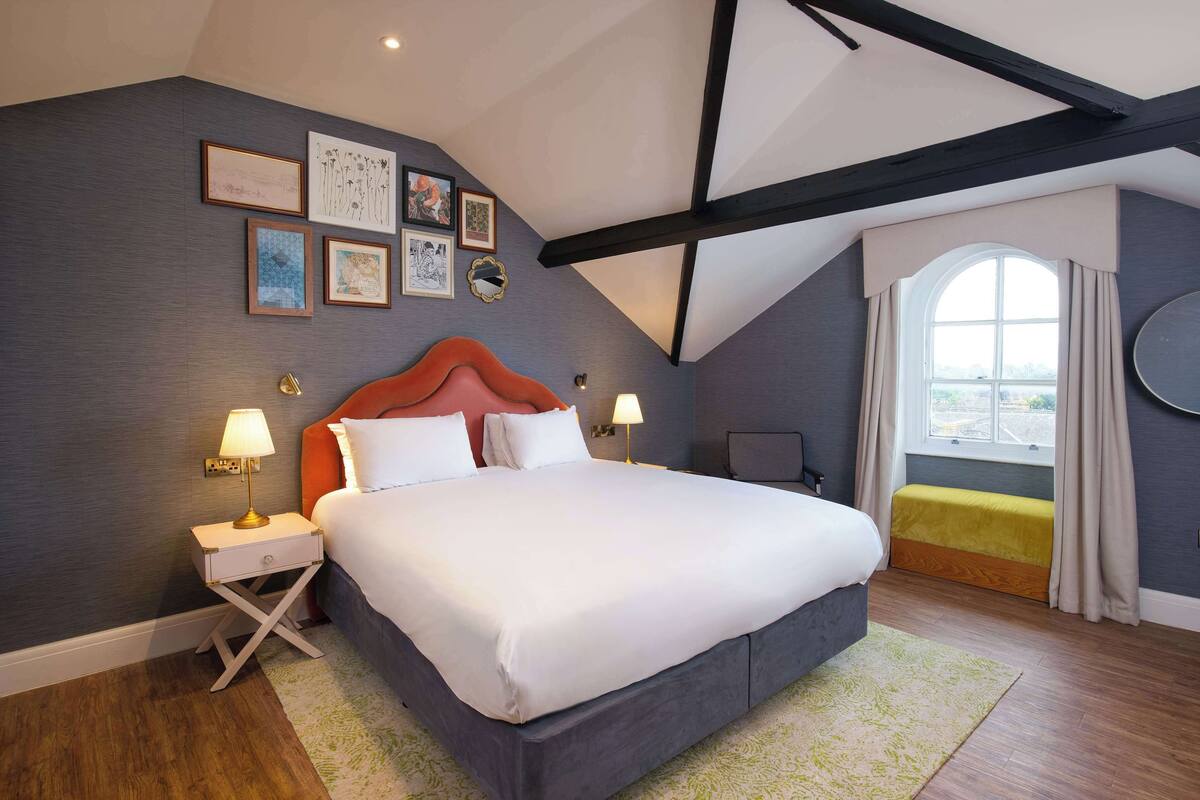 Quarto com uma cama grande, mesa de cabeceira com luminária, janela com cortinas e obras de arte na parede.