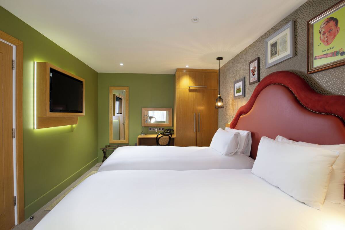 Quarto de hotel com duas camas, uma escrivaninha com cadeira, espelho e televisão na parede.