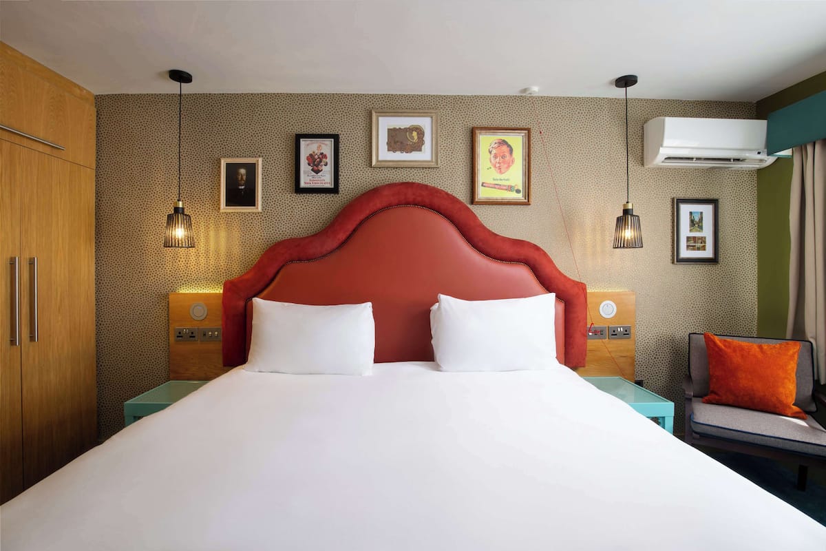 Quarto de hotel com uma cama grande, duas mesas de cabeceira, cabeceira estofada vermelha e obras de arte penduradas na parede.