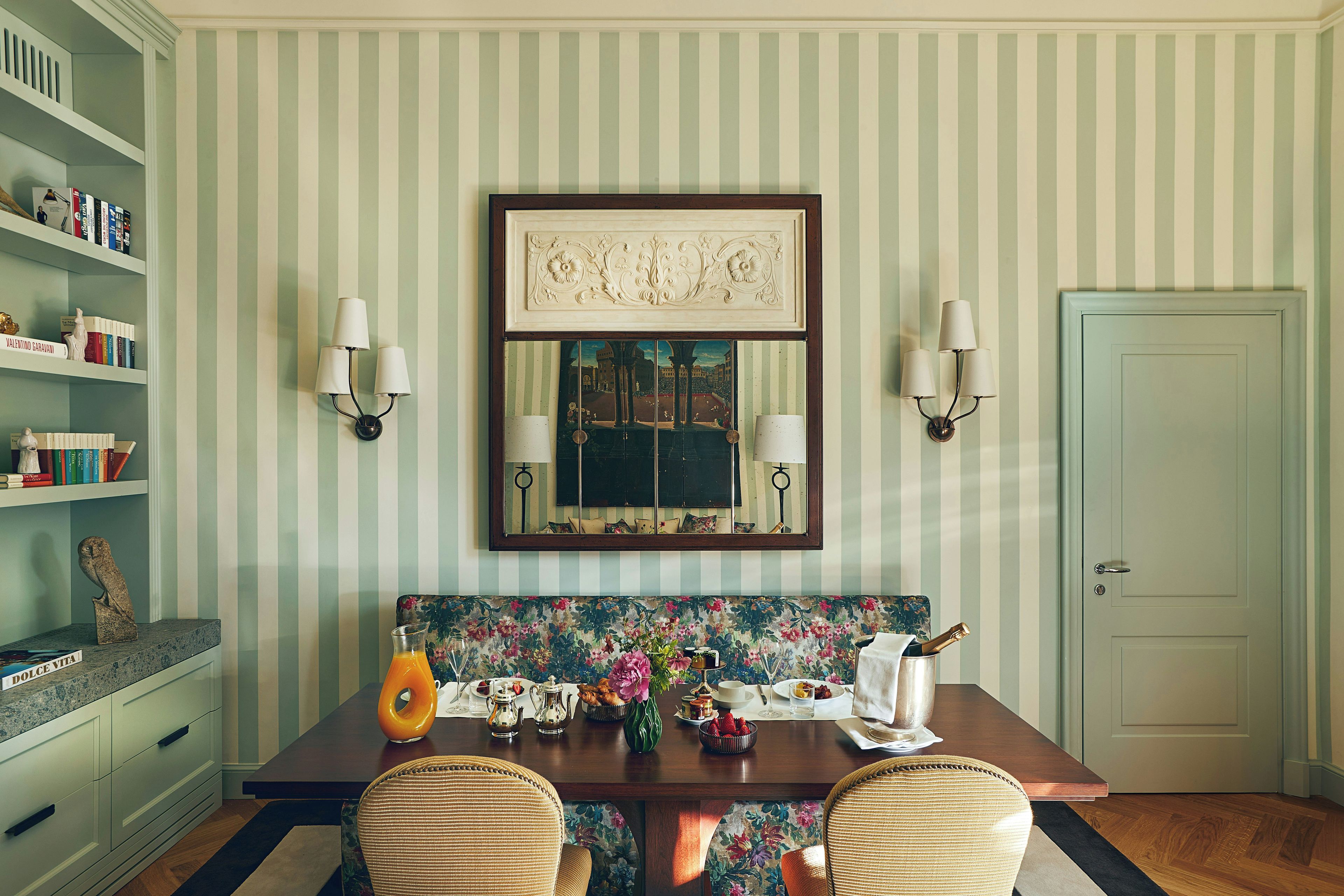 Uma sala de jantar com papel de parede listrado, uma mesa de madeira, duas cadeiras, um banco floral e uma estante de livros.
