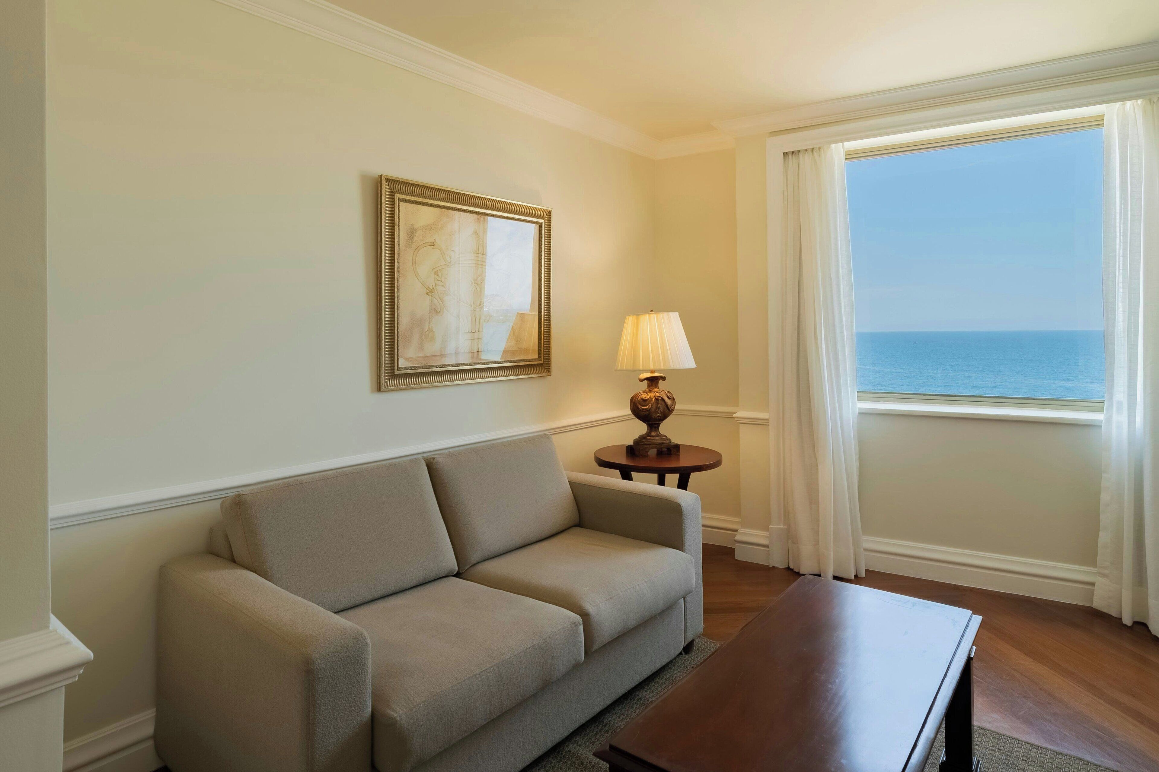 Quarto com sofá bege, mesa de centro de madeira, luminária, obra de arte emoldurada e vista para o mar através de uma janela com cortinas brancas.