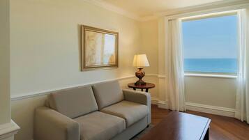 Quarto com sofá bege, mesa de centro de madeira, luminária, obra de arte emoldurada e vista para o mar através de uma janela com cortinas brancas.