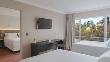 Quarto de hotel com cama, escrivaninha com televisão e janela com vista para árvores.