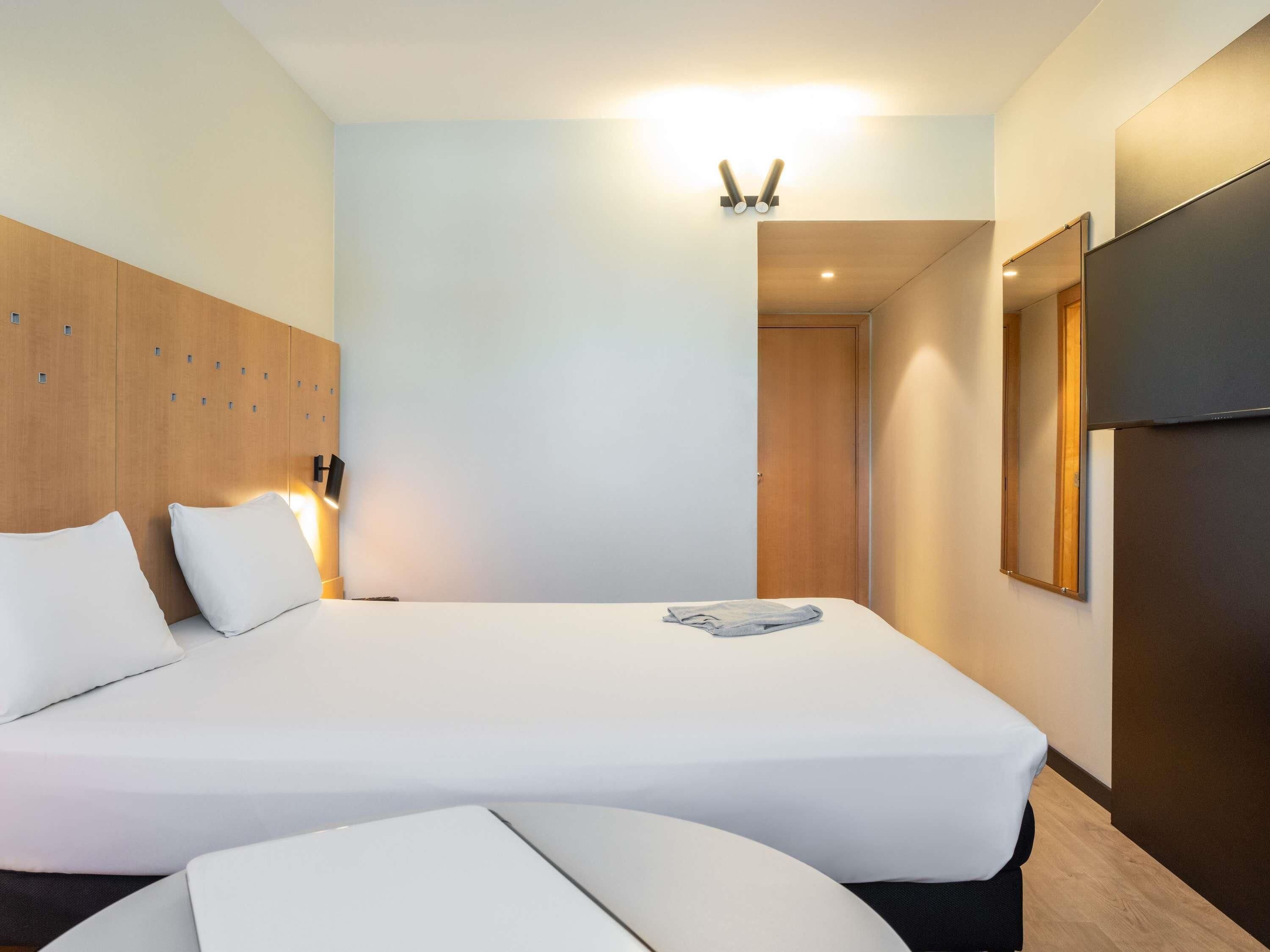 Quarto de hotel com uma cama grande, uma mesinha de cabeceira, uma luminária na parede e uma porta que dá para outro cômodo.