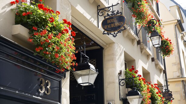 Entrada de um hotel com uma placa indicando "HOTEL D'AUBUSSON", uma porta preta com o número "33" e caixas de flores com flores vermelhas.