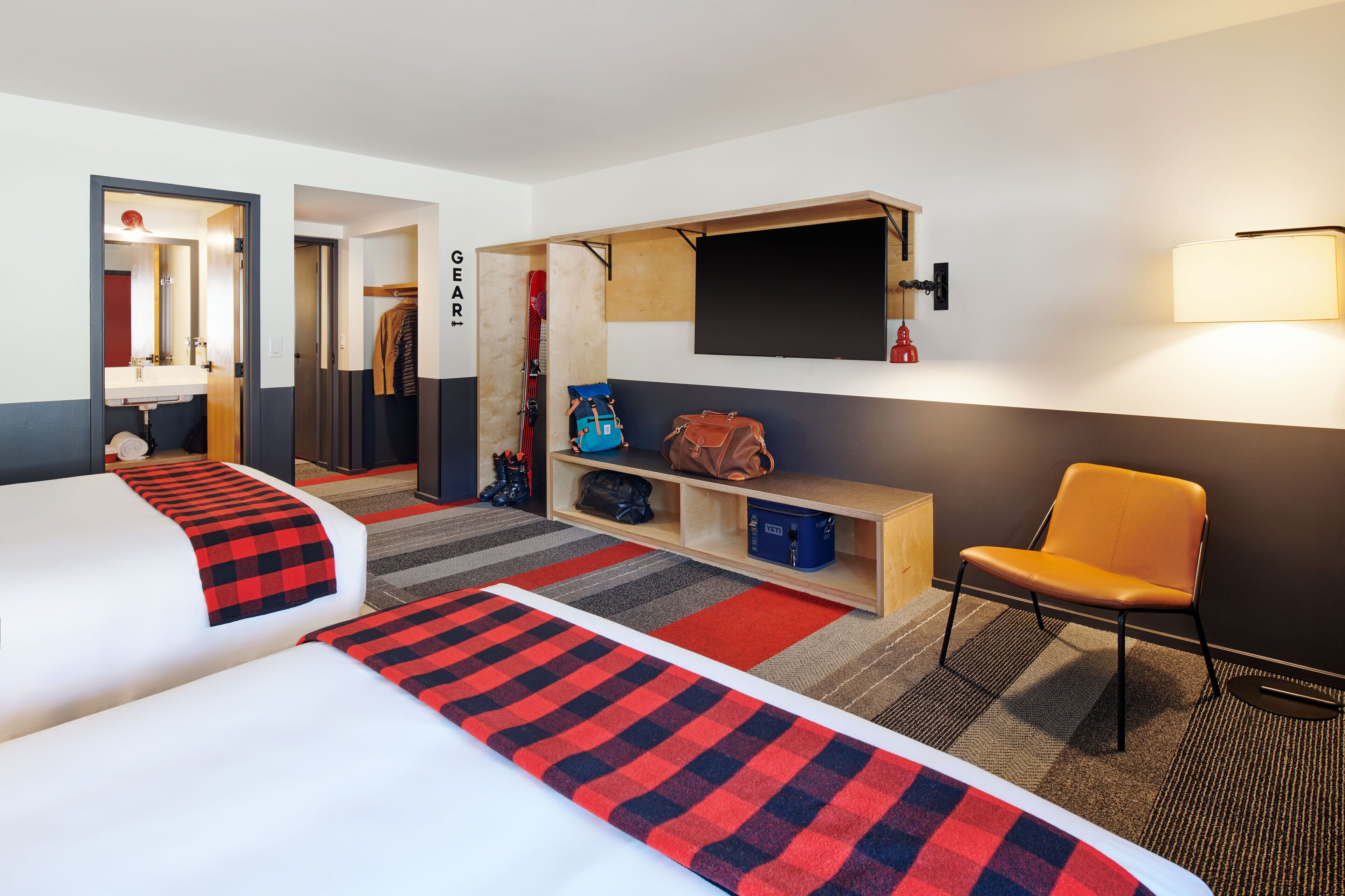Quarto de hotel com duas camas, uma televisão na parede, uma prateleira de madeira com malas e uma cadeira.