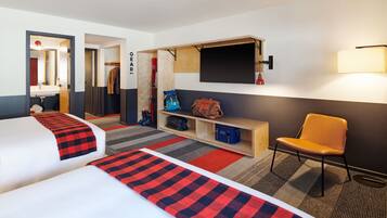 Quarto de hotel com duas camas, uma televisão na parede, uma prateleira de madeira com malas e uma cadeira.