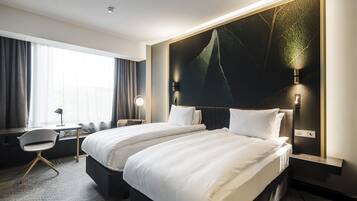 Quarto de hotel com duas camas, uma escrivaninha, uma cadeira e uma janela grande com cortinas.