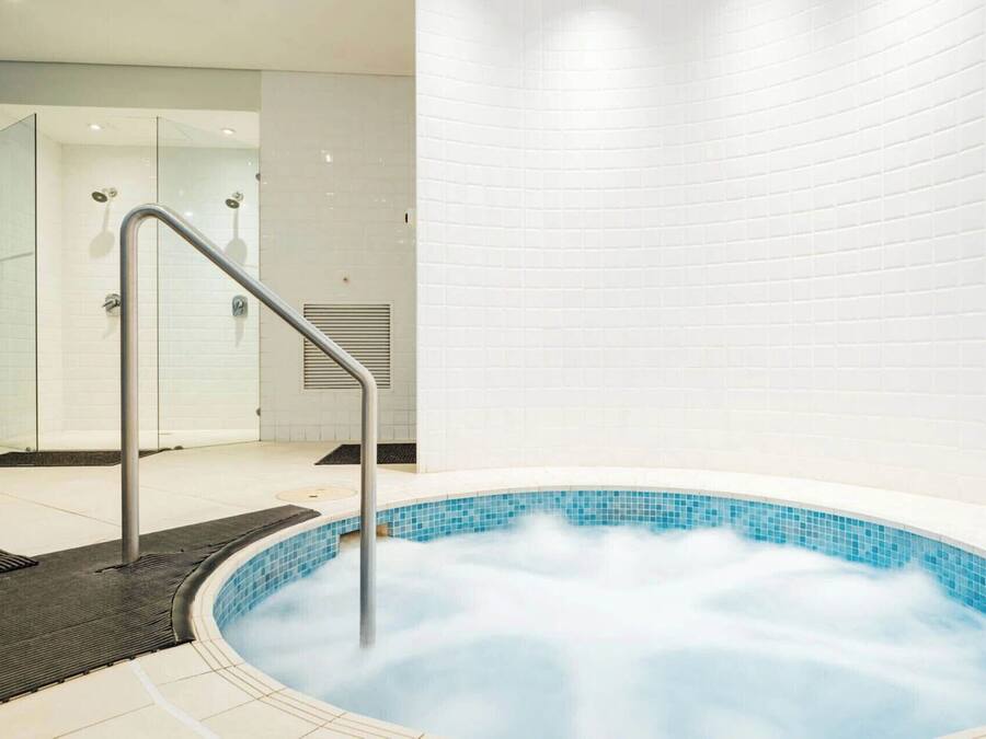 Área de spa com hidromassagem, cercada por paredes de azulejos brancos e entrada por porta de vidro.
