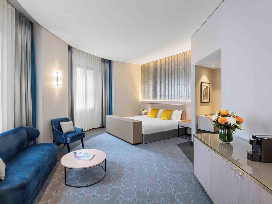 Quarto de hotel com cama, sofá azul, mesa pequena, escrivaninha e um vaso de flores.