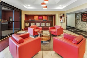 Um hall de entrada com sofás de couro vermelho, uma mesa de centro de vidro e uma mesa de recepção com a placa 'Wi-Fi Gratuito em Todos os Quartos'.