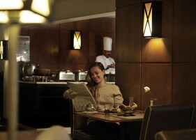 Uma mulher lendo um cardápio em uma mesa de restaurante, com um chef ao fundo.