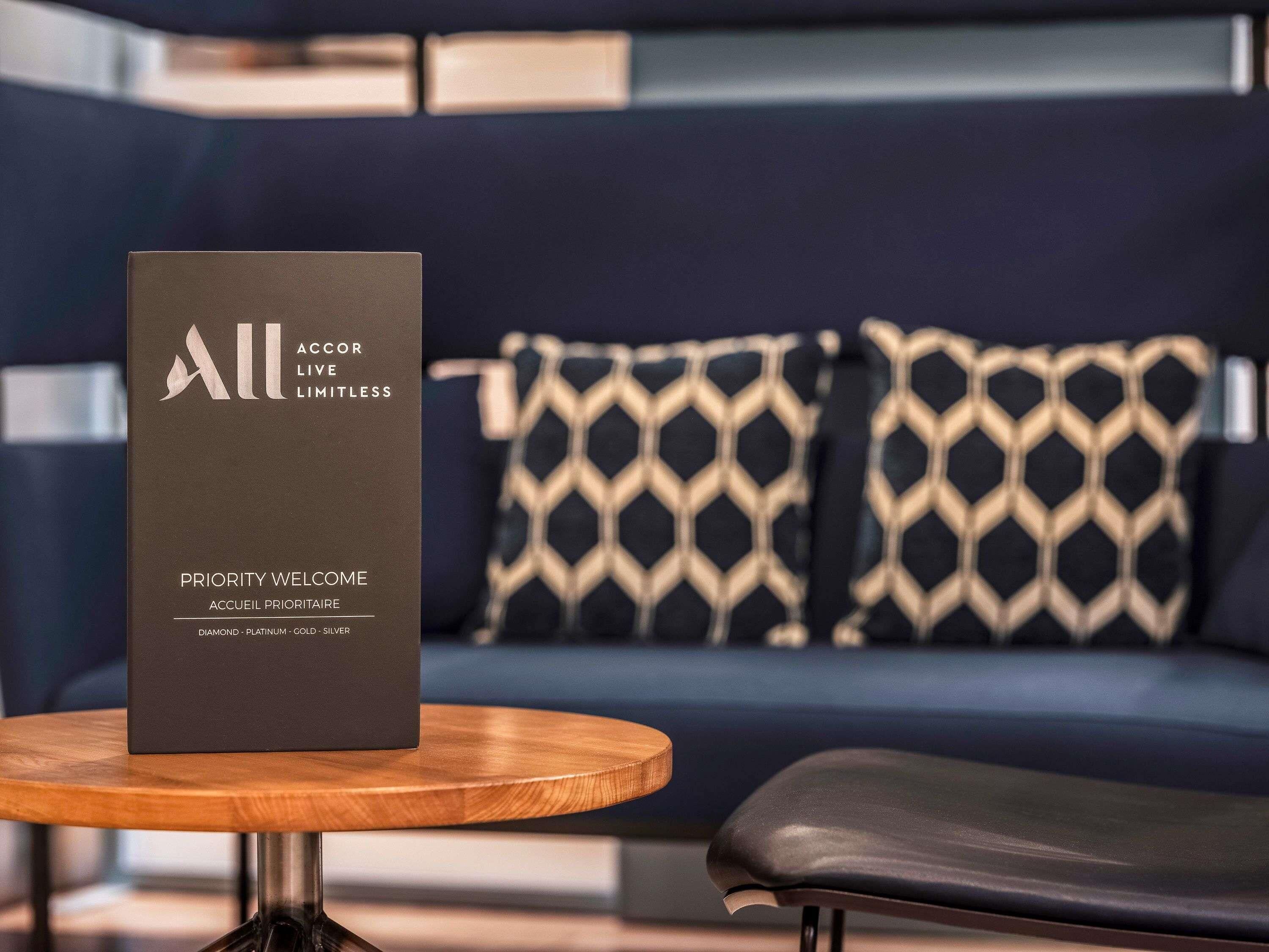 Placa de boas-vindas prioritária com os dizeres 'All Accor Live Limitless' e 'Priority Welcome', colocada sobre uma mesa de madeira em frente a um sofá azul escuro com almofadas estampadas.
