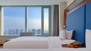 Quarto de hotel com duas camas, vista para a cidade, escrivaninha com telefone e prato de frutas.