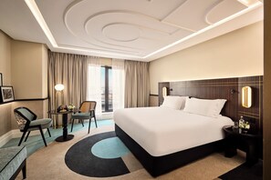 Quarto de hotel com uma cama grande, uma escrivaninha com cadeira, mesa de cabeceira e vista para a cidade pela janela.