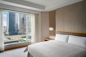 Quarto de hotel com uma cama grande, mesa de cabeceira, luminária e vista para prédios altos através de uma janela ampla.