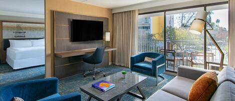 Quarto de hotel com uma cama grande, um sofá, duas poltronas, uma mesa de centro, uma TV fixada na parede e uma varanda com vista para a cidade.