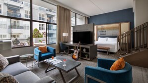 Quarto de hotel moderno com uma TV grande, assentos confortáveis, uma guitarra e vista para a cidade.