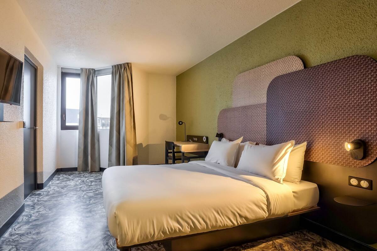 Quarto de hotel com uma cama grande, uma escrivaninha e vista para a cidade pela janela.