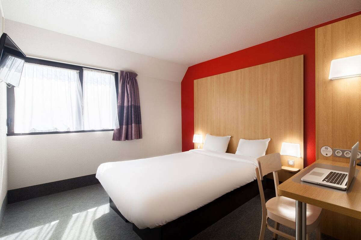 Quarto de hotel com cama, escrivaninha com laptop, cadeira, janela com cortinas e uma parede de destaque vermelha.